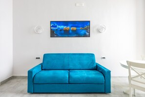 Quarto com sofá azul, pintura de uma paisagem costeira e mesa de jantar branca com cadeiras.