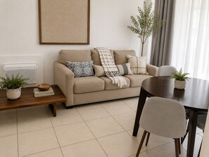 Uma sala de estar moderna com um sofá bege, uma mesa de centro e uma mesa de jantar com cadeiras. Há plantas em vasos na mesa de centro e um quadro grande na parede.