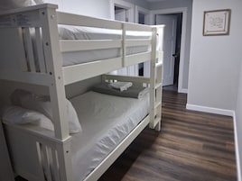 Uma cama beliche com uma cama de baixo e uma cama individual, piso de madeira, uma porta e um quadro pendurado na parede.