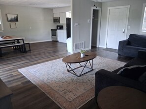 Sala de estar com sofá escuro, mesa de centro com planta, tapete e área de jantar com banco e mesa.