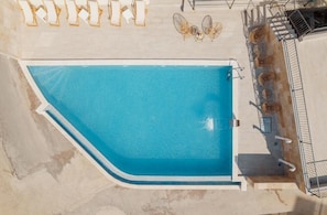 Vista aérea de uma piscina com borda curva, rodeada por um deck revestido com azulejos e algumas cadeiras de descanso.