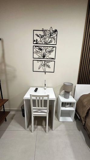 Quarto pequeno e bem arrumado, com escrivaninha branca, cadeira e mesa de cabeceira. Um controle remoto está sobre a escrivaninha, e uma luminária sobre a mesa de cabeceira.