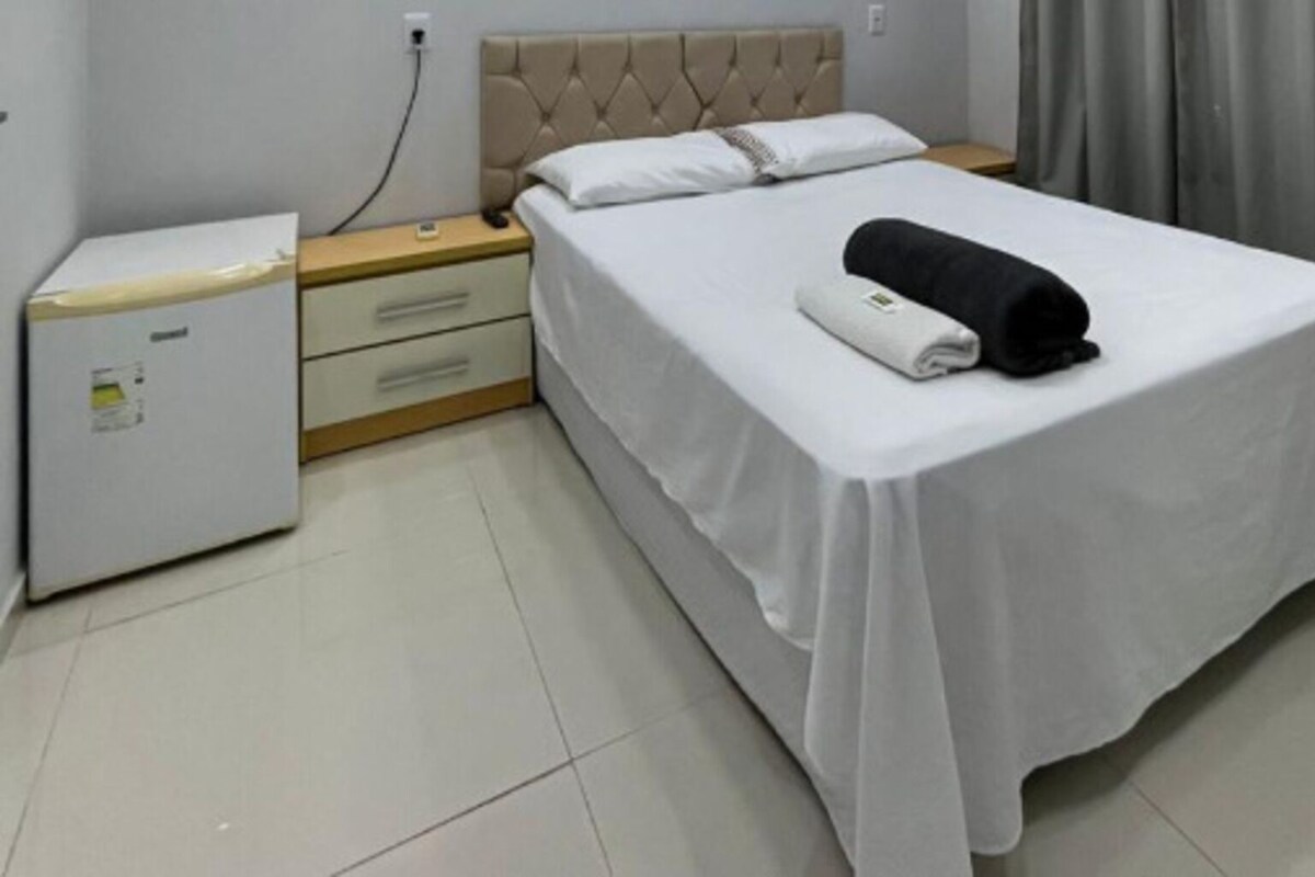 Quarto de hotel com cama, mesa de cabeceira, mini-geladeira e tomada.