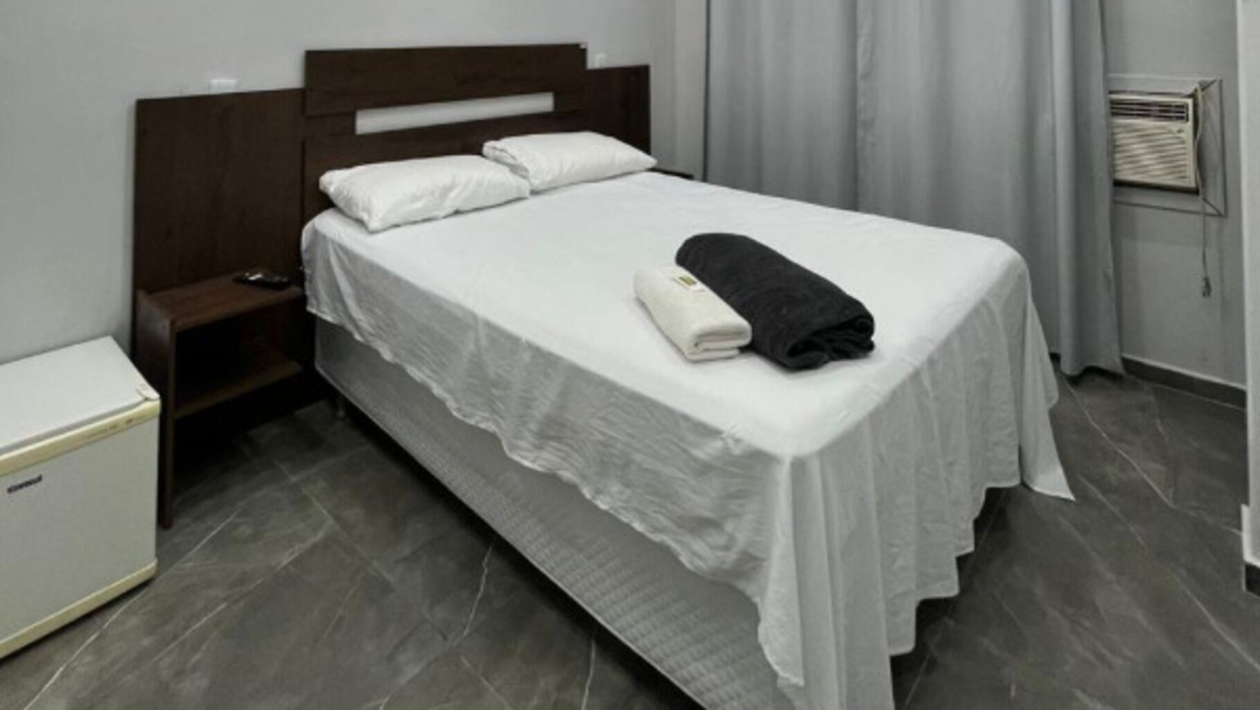 Uma cama de solteiro com um cobreleito branco, duas almofadas, uma toalha preta e uma mesa de cabeceira com abajur.
