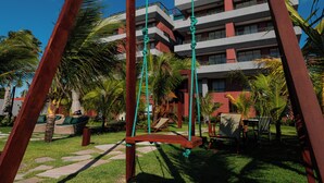 Um balanço com estrutura de madeira e cordas cor turquesa, ao fundo um edifício de vários andares com varandas e palmeiras.