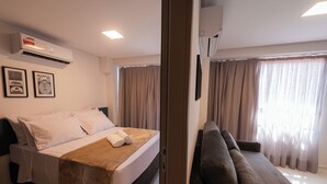 Quarto de hotel com cama, sofá, janela com cortinas e ar condicionado na parede.