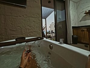 Uma pessoa relaxando em uma banheira de hidromassagem com um copo de vinho, uma janela e vista para prédios.