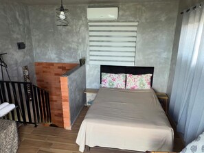 Quarto com cama, mesa de cabeceira, pequena mesa, divisória de tijolos, radiador e janela com persianas.
