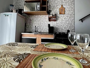 Cozinha equipada com frigorífico, micro-ondas e máquina de café. Há uma mesa de jantar com pratos e copos.