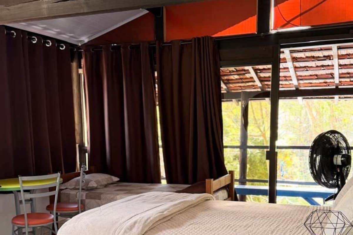 Quarto com cama, escrivaninha, cadeira, ventilador e janela com cortinas.