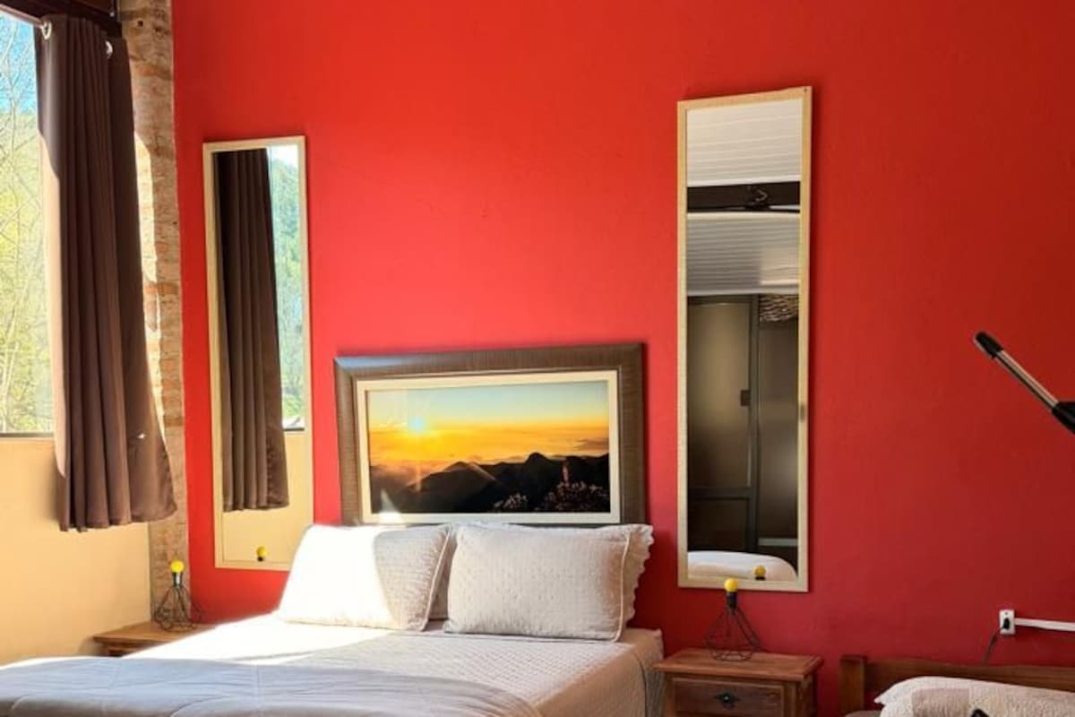 Quarto com parede vermelha, duas camas, um espelho grande e uma janela com cortinas.