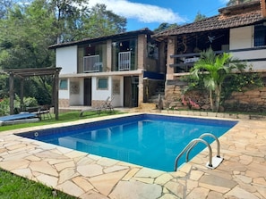 Piscina com azulejos de pedra, um edifício de dois andares com varanda e um pergolado.