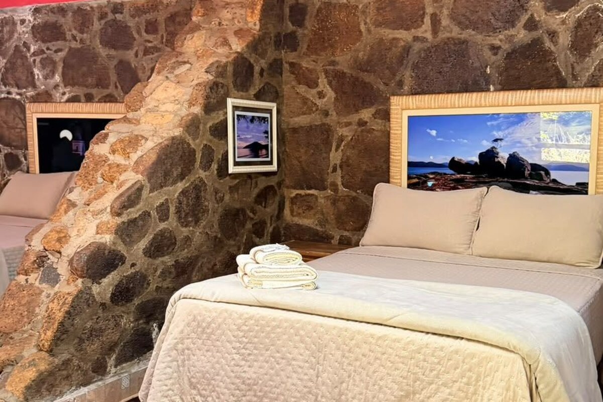 Quarto com parede de pedra, cama com colcha bege, tapete escuro e quadro na parede.