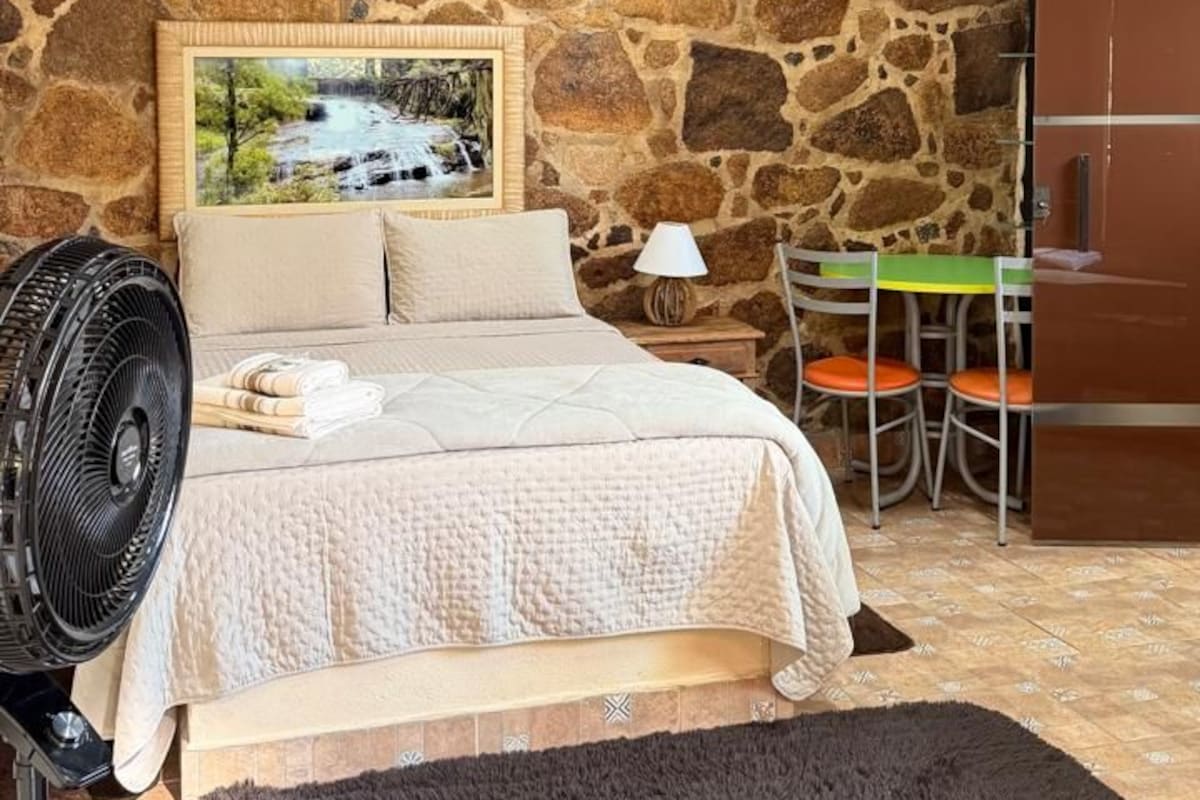Quarto com parede de pedra, uma cama com colcha bege, um ventilador, uma mesa com cadeiras e vista para um rio.