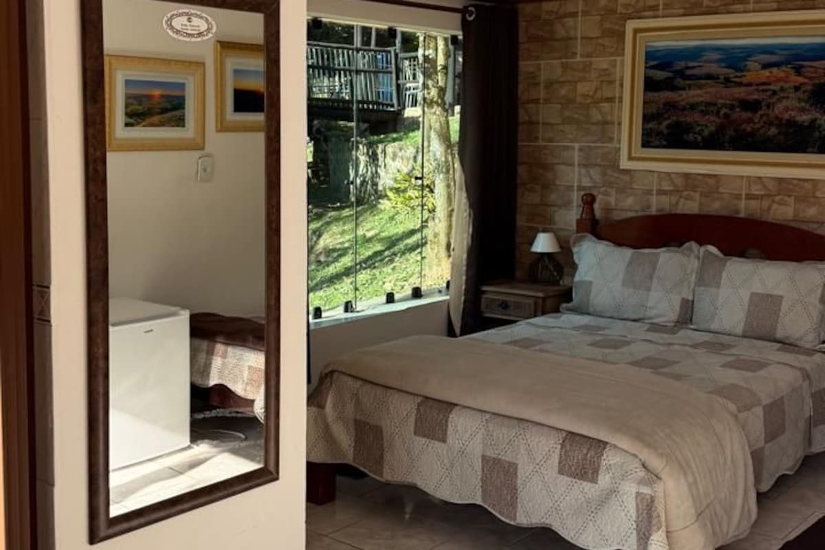 Quarto com cama, mesa de cabeceira, janela com vista para o verde e um quadro na parede.
