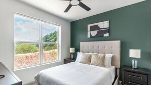 Um quarto com uma cama, duas mesas de cabeceira com luminárias, um ventilador de teto, uma janela grande com vista e uma obra de arte abstrata na parede.
