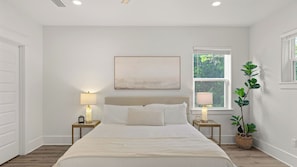 Quarto com cama, duas mesas de cabeceira com abajures, janela com vista para vegetação e pintura abstrata pendurada na parede.