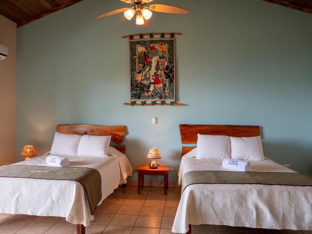 Quarto de hotel com duas camas, um ventilador de teto, tapeçaria na parede e abajures.