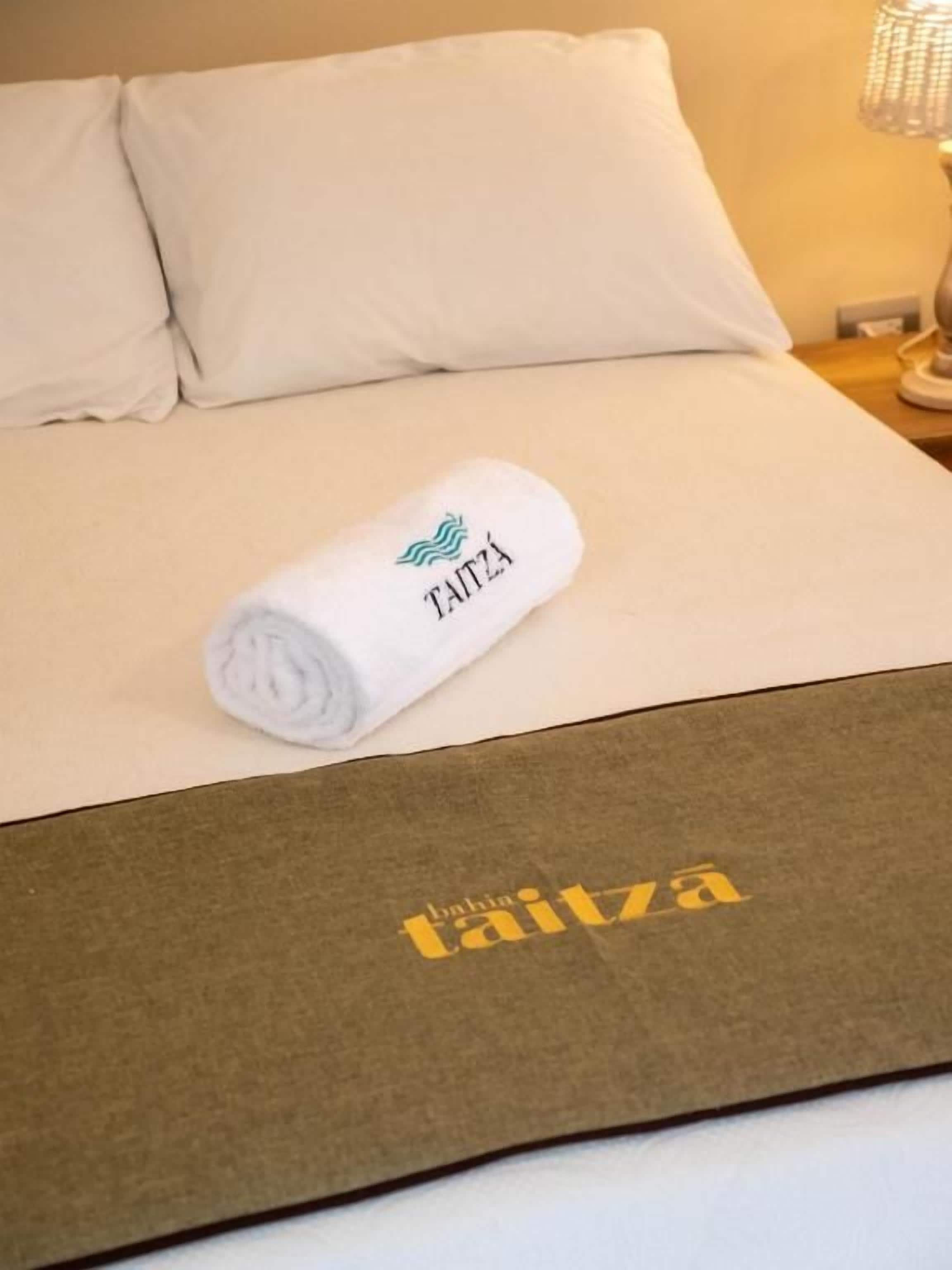 Cama bem arrumada com toalha enrolada e manta marrom com a marca 'Taitza' visível.