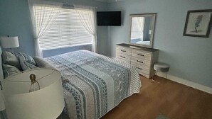Quarto com cama, ventoinha no teto, televisão, cômoda, espelho e janela com persianas.