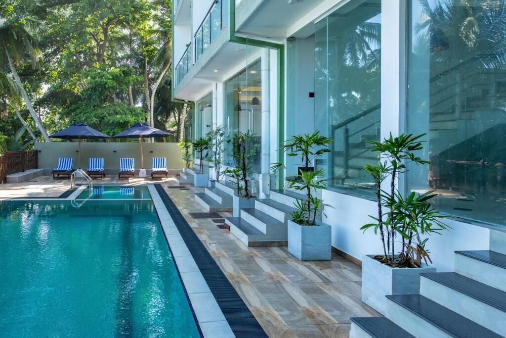 Piscina com espreguiçadeiras e guarda-sóis, rodeada por plantas em vasos e um edifício moderno com grandes janelas.