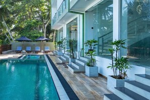 Piscina com espreguiçadeiras e guarda-sóis, rodeada por plantas em vasos e um edifício moderno com grandes janelas.
