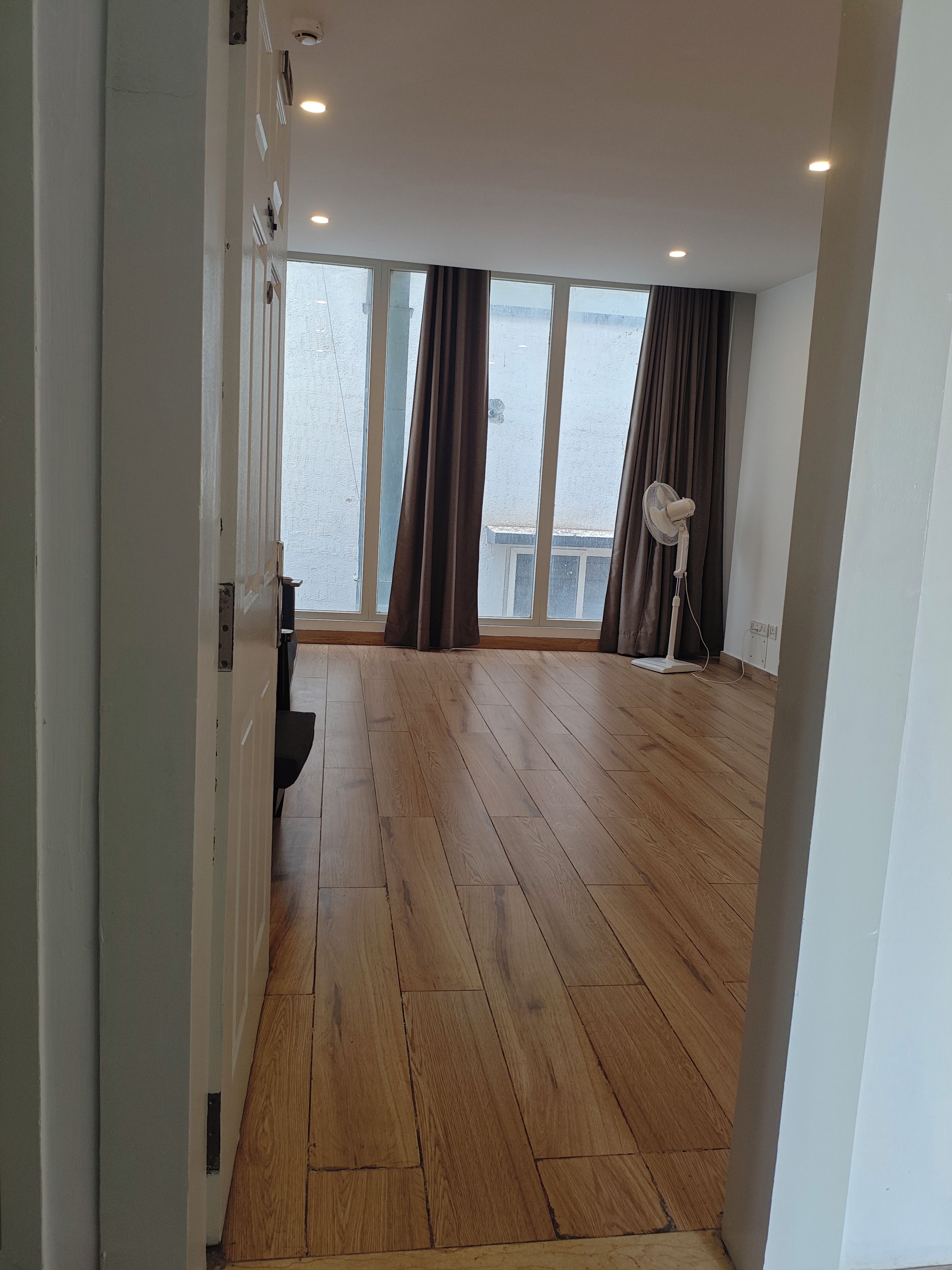 Quarto com piso de madeira, janelas amplas com cortinas e um ventilador.