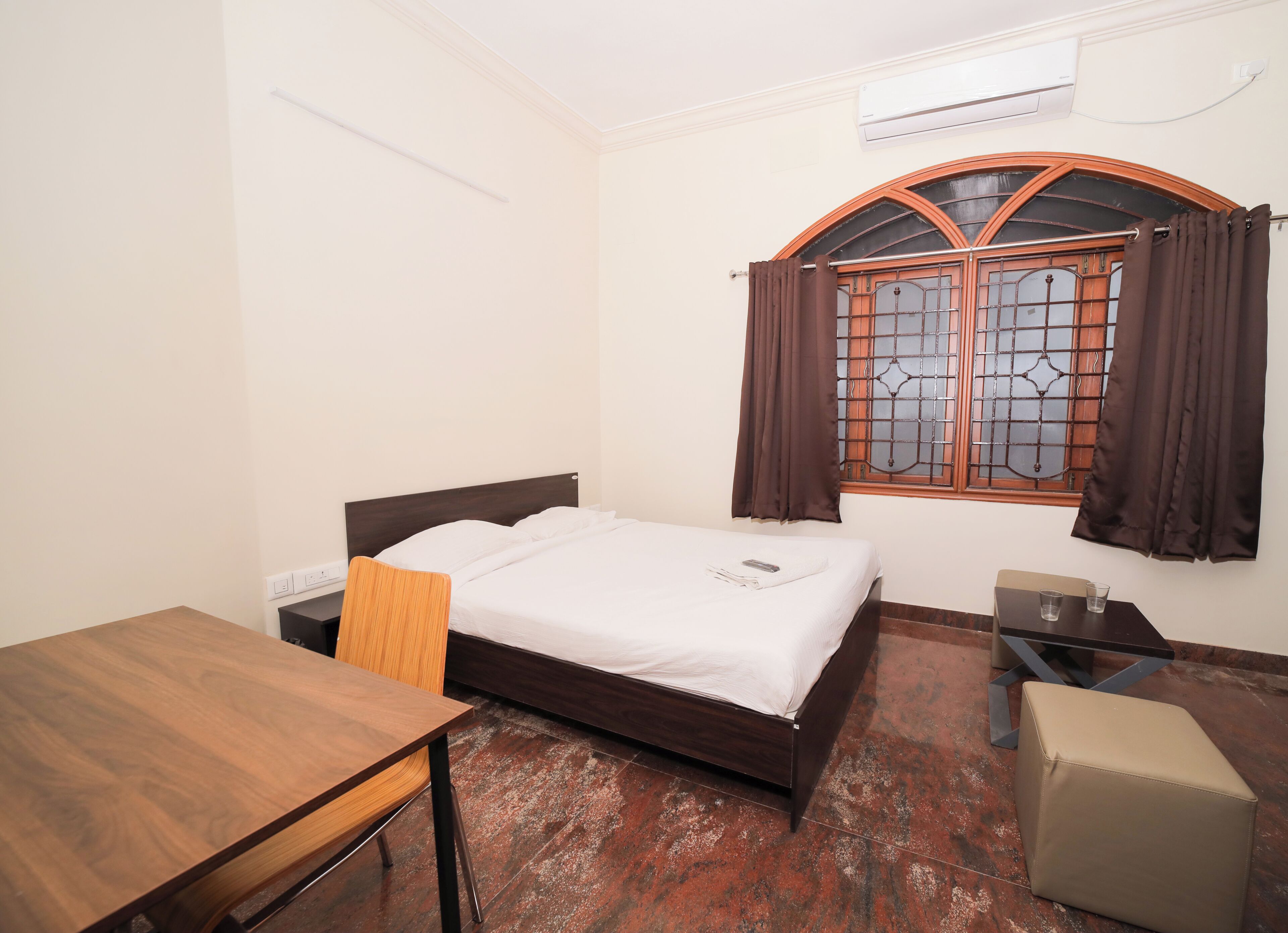 Quarto com cama, mesa de madeira, cadeira, pequena mesa com copos e janela com cortinas.