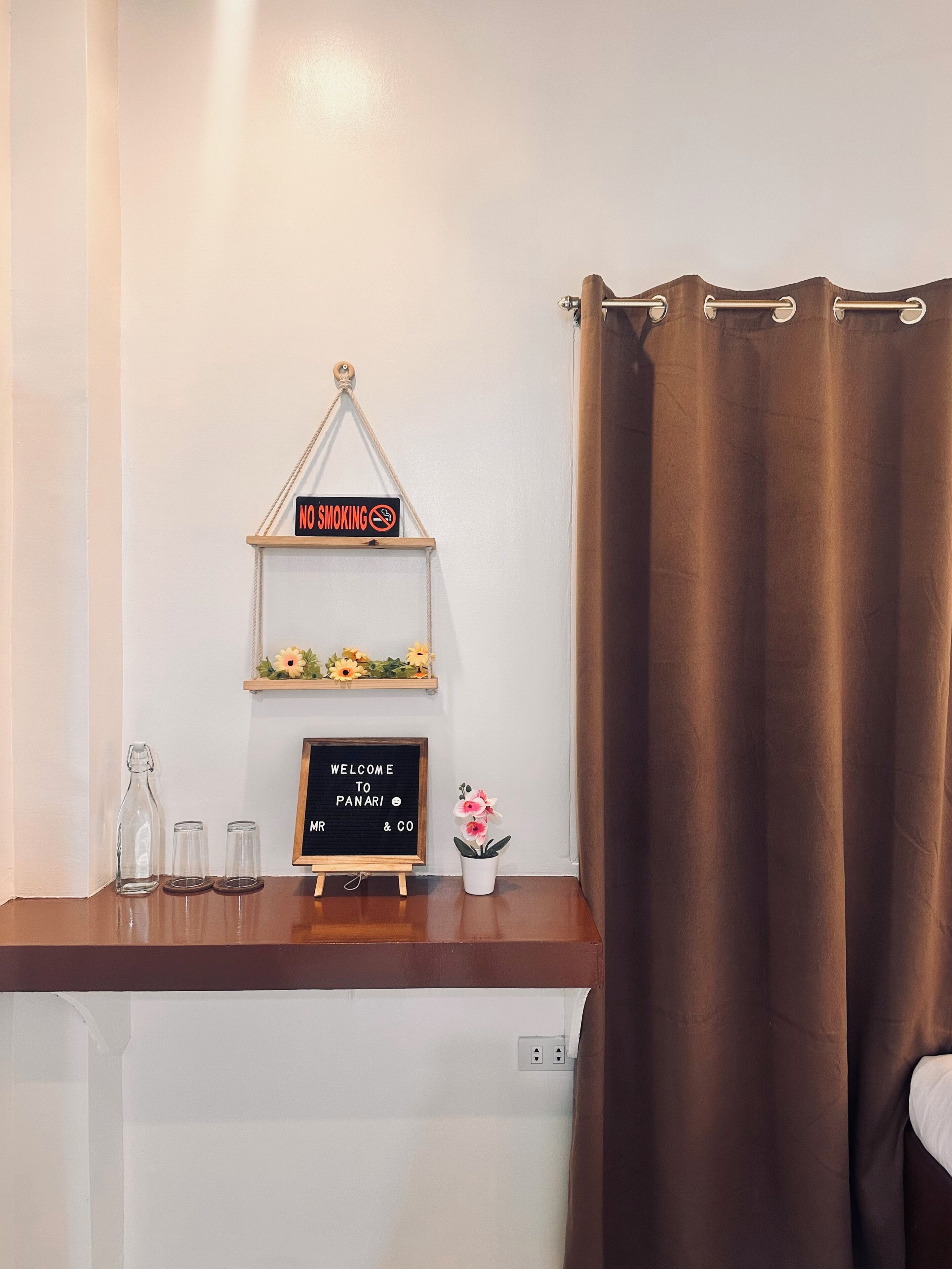 Quarto com uma estante de madeira exibindo um aviso 'Não Fumar', uma placa de boas-vindas e um pequeno vaso de planta.