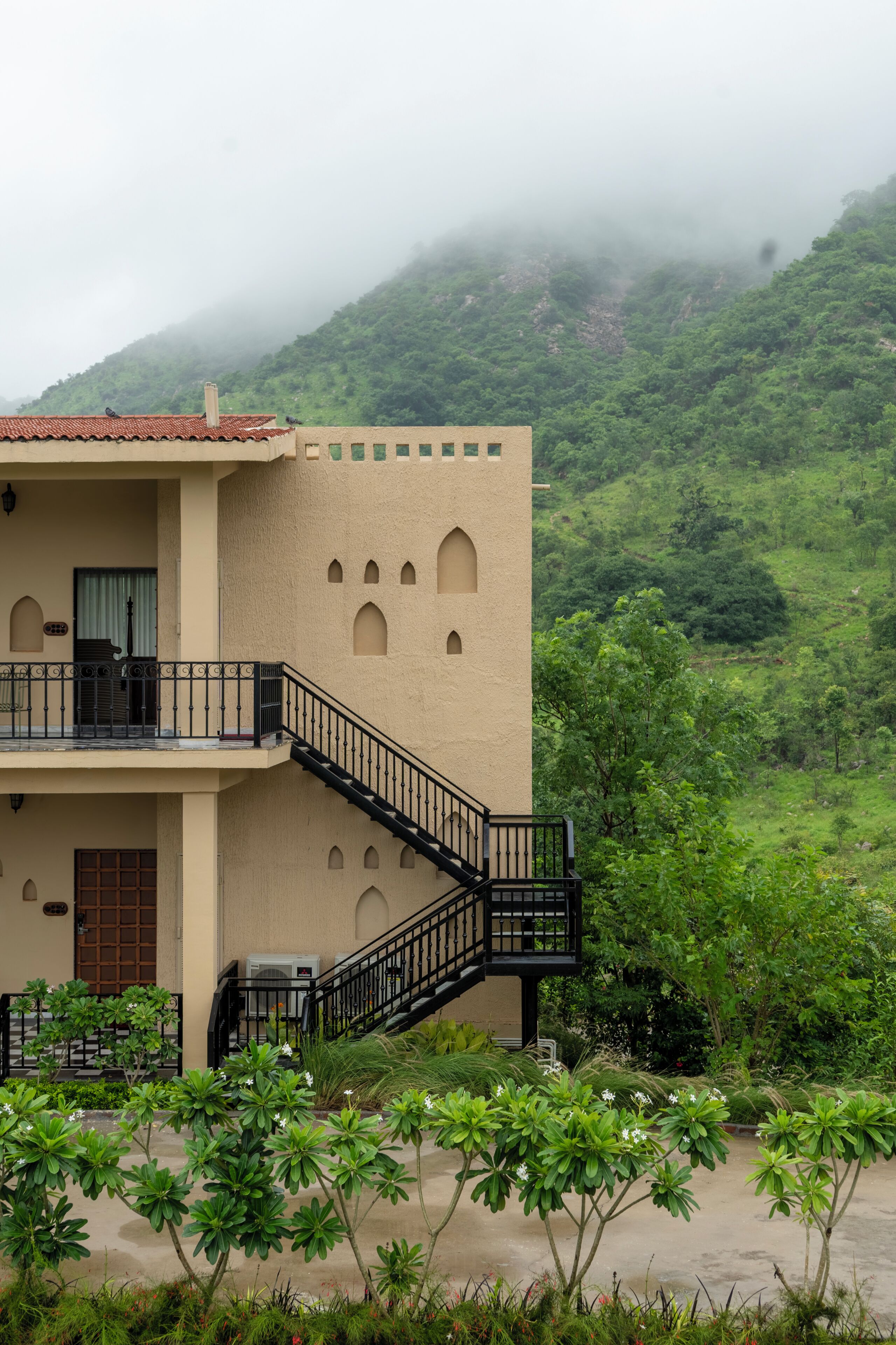 Um edifício com escadaria que dá para uma varanda, rodeado por vegetação exuberante e uma paisagem montanhosa.