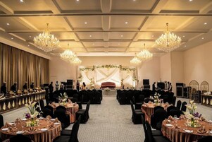 Salão de festas com mesas redondas, cadeiras pretas e um palco com decorações florais.