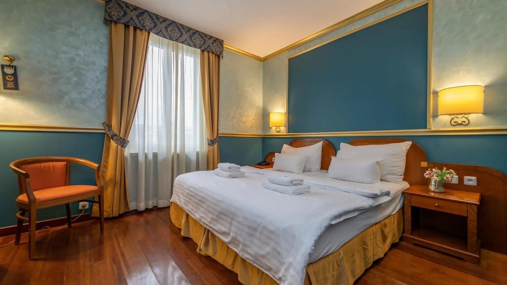 Quarto de hotel com cama, cadeira, mesa de cabeceira e janela com cortinas.