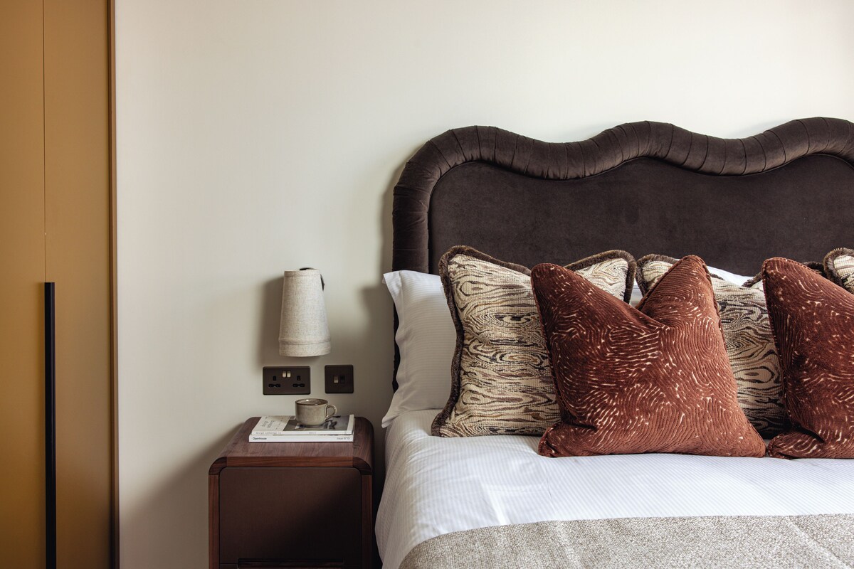 Cama bem arrumada com cabeceira escura, mesa de cabeceira com abajur e tomada.