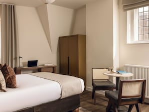 Quarto de hotel com cama, escrivaninha, cadeira, radiador e janela.