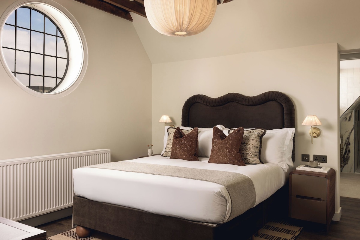 Quarto com cama grande, janela redonda e vigamento de madeira no teto.