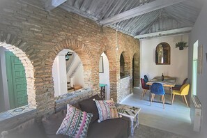 Quarto com paredes de tijolo aparente, área de jantar com mesa e cadeiras, e sofá com almofadas estampadas.