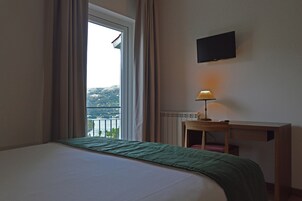 Quarto de hotel com uma cama, uma escrivaninha com luminária, televisão na parede e vista para a água através da janela.