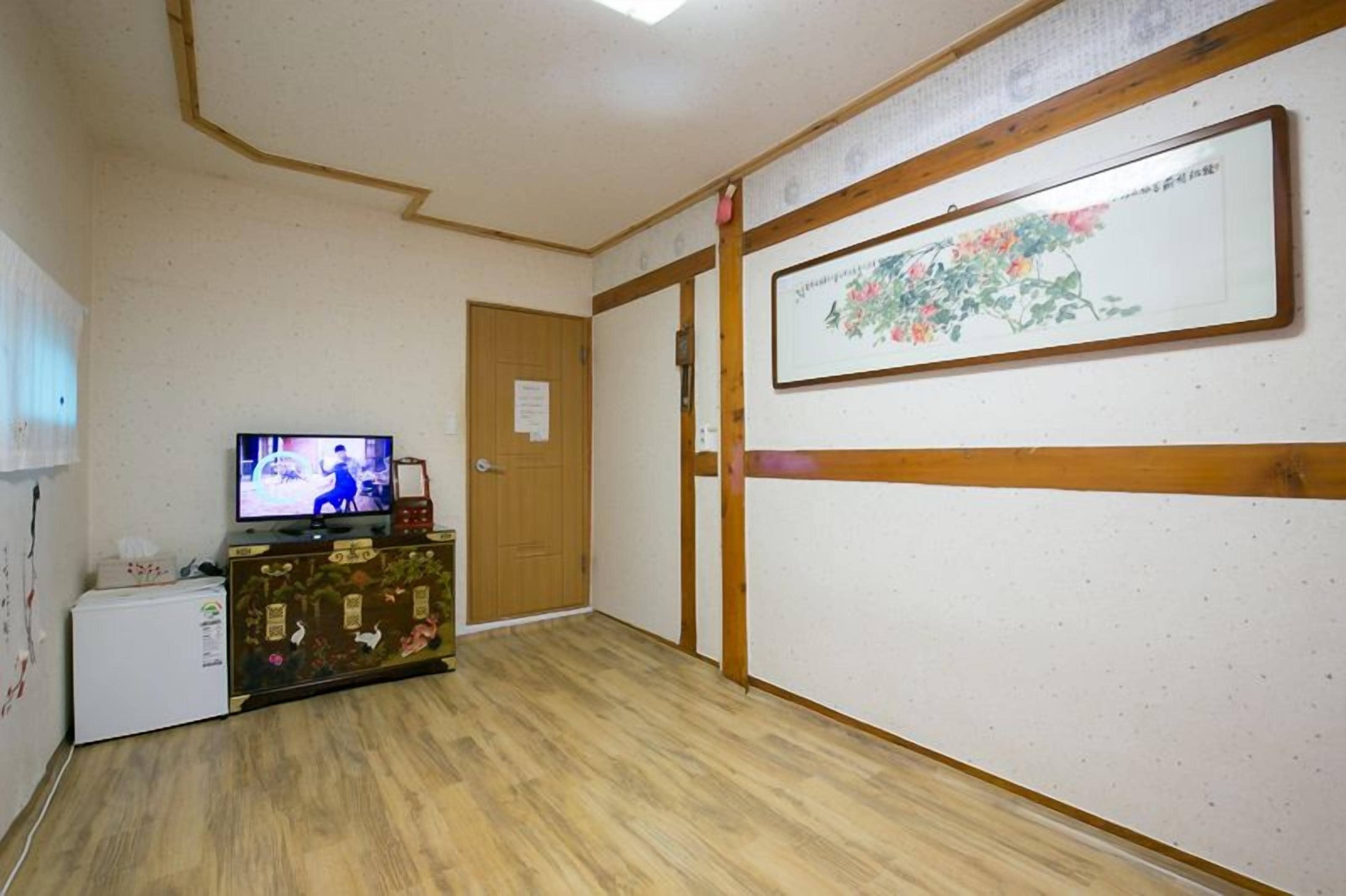 Quarto com piso de madeira, televisão sobre um móvel, porta e quadro pendurado na parede.