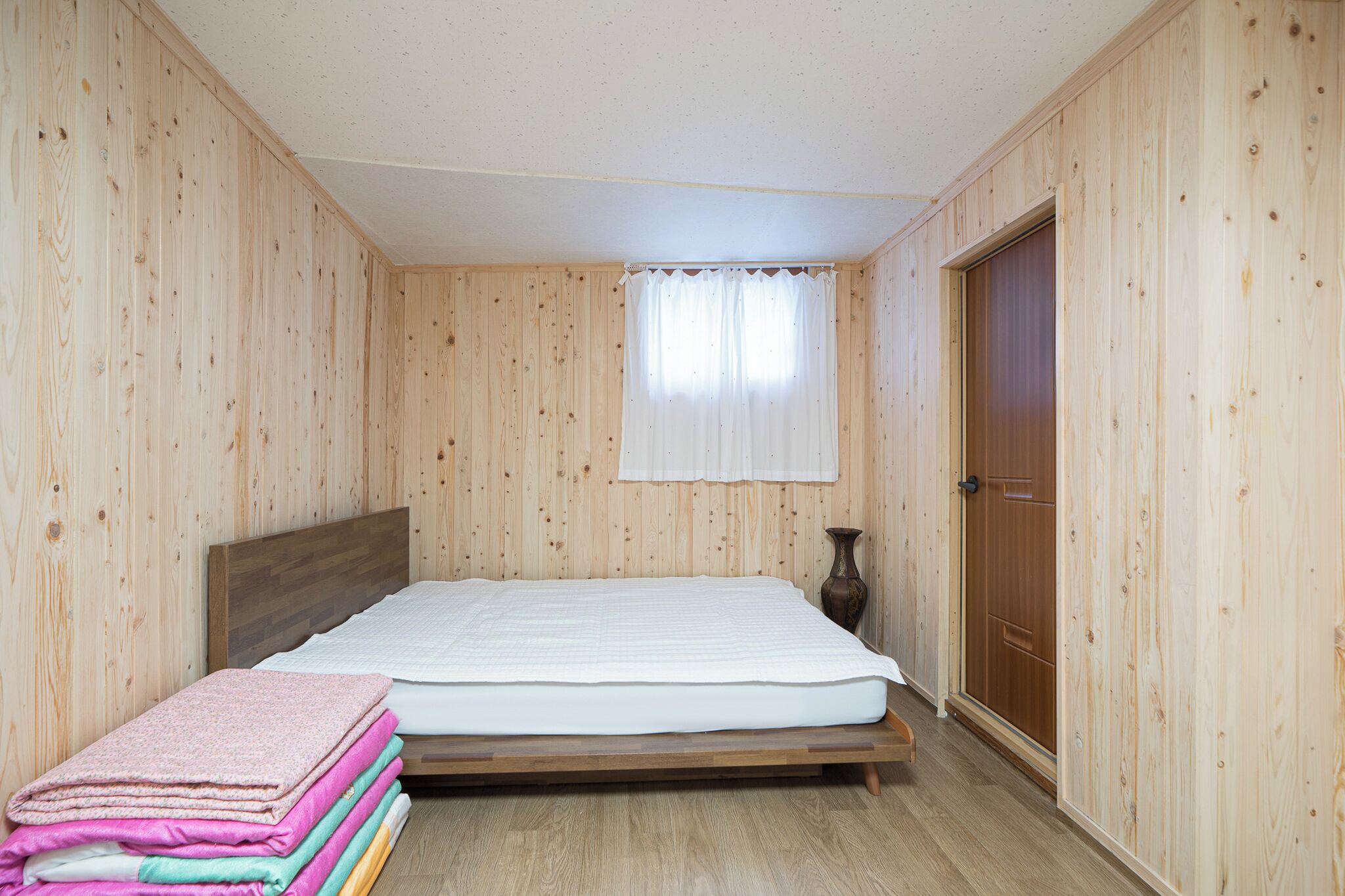 Quarto com paredes de madeira, cama com colchão branco, cabeceira de madeira, janela com cortinas brancas e porta de madeira.
