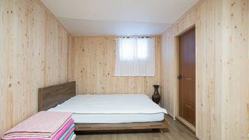 Quarto com paredes de madeira, cama com colchão branco, cabeceira de madeira, janela com cortinas brancas e porta de madeira.