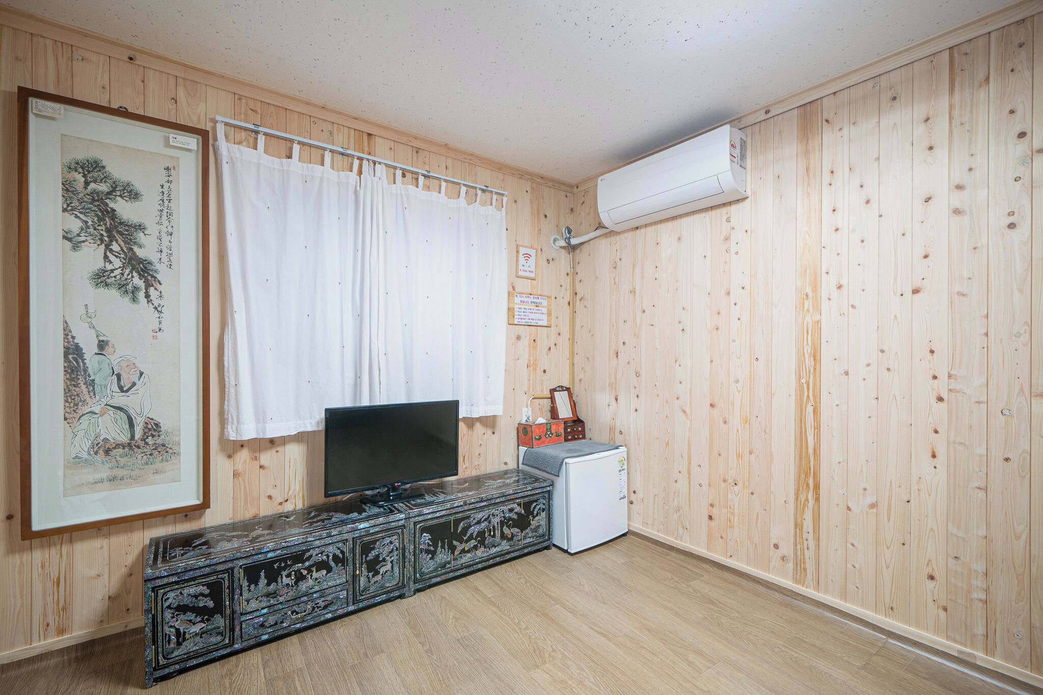 Quarto com paredes de madeira, televisão de tela plana sobre um móvel decorativo, ar condicionado branco e uma obra de arte emoldurada.