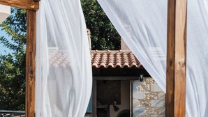 Uma pérgula de madeira com cortinas brancas, um edifício de pedra com telhado em telhas e um chão ladrilhado.