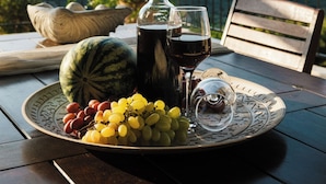 Uma mesa com uma garrafa, copos, uvas e melancia em um terraço com vista para as montanhas.