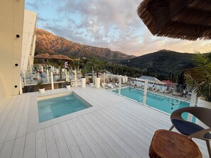 Área de piscina com vista para as montanhas, cadeiras de descanso e estrutura com telhado de palha.