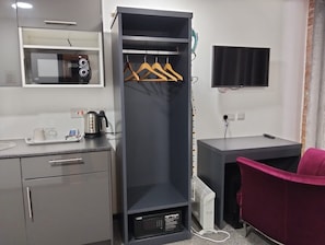 Quarto de hotel compacto com cozinha compacta, uma secretária pequena, televisão e guarda-roupa com cabides.