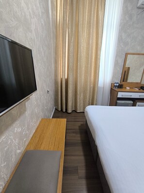 Quarto de hotel com cama, televisão na parede, mesa de cabeceira de madeira e espelho.