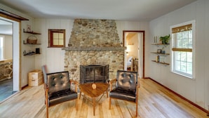Quarto com lareira de pedra, duas poltronas de couro, uma mesa de madeira e janela com persianas.