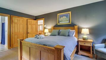 Quarto com cama de madeira, duas mesas de cabeceira com abajures, um quadro na parede e uma porta que dá para outro cômodo.
