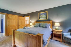 Quarto com cama de madeira, duas mesas de cabeceira com abajures, um quadro na parede e uma porta que dá para outro cômodo.