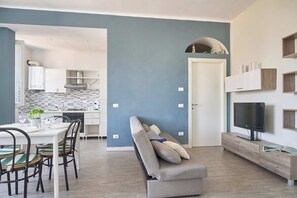 Uma sala de estar moderna com um sofá cinzento, uma área de jantar com uma mesa e cadeiras brancas, uma cozinha com armários brancos e um painel de azulejos, e uma TV sobre um suporte de madeira.